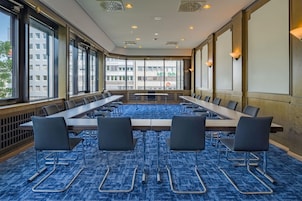 Sala de conferência com uma mesa comprida e cadeiras, janelas amplas e carpete azul.