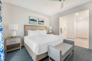 Quarto de hotel com uma cama grande, duas mesas de cabeceira com luminárias, uma cadeira e uma porta que dá para outro cômodo.