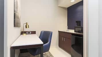 Quarto de hotel compacto com uma pequena escrivaninha, uma cadeira azul, cafeteira e micro-ondas.