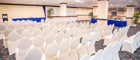 Uma sala de conferências com fileiras de cadeiras brancas, uma mesa azul com arranjos florais e um pódio com a inscrição 'PEGAUSUS'.
