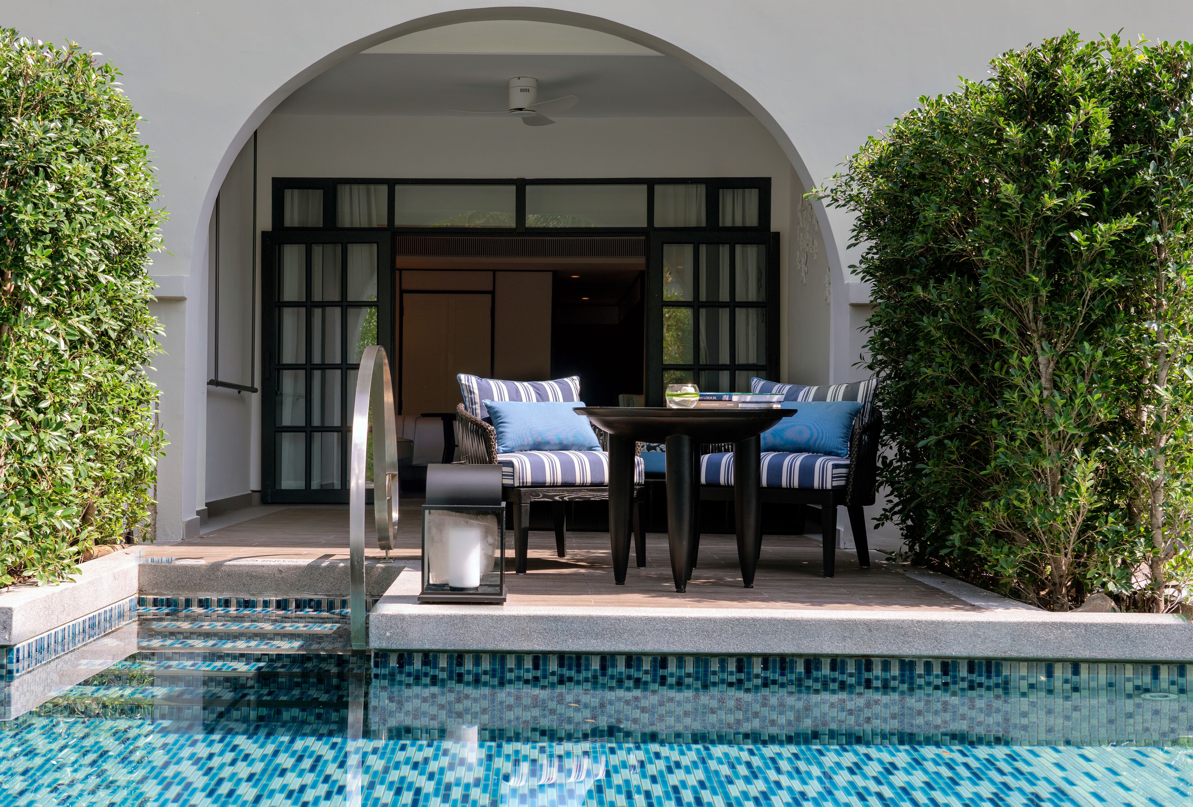 Uma área de piscina externa com piso de azulejos, duas espreguiçadeiras com almofadas listradas, uma mesa e um ventilador de teto.