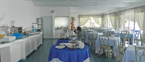 Uma ampla área de refeições com mesas postas, um buffet com diversos pratos e vista para o exterior através de grandes janelas.