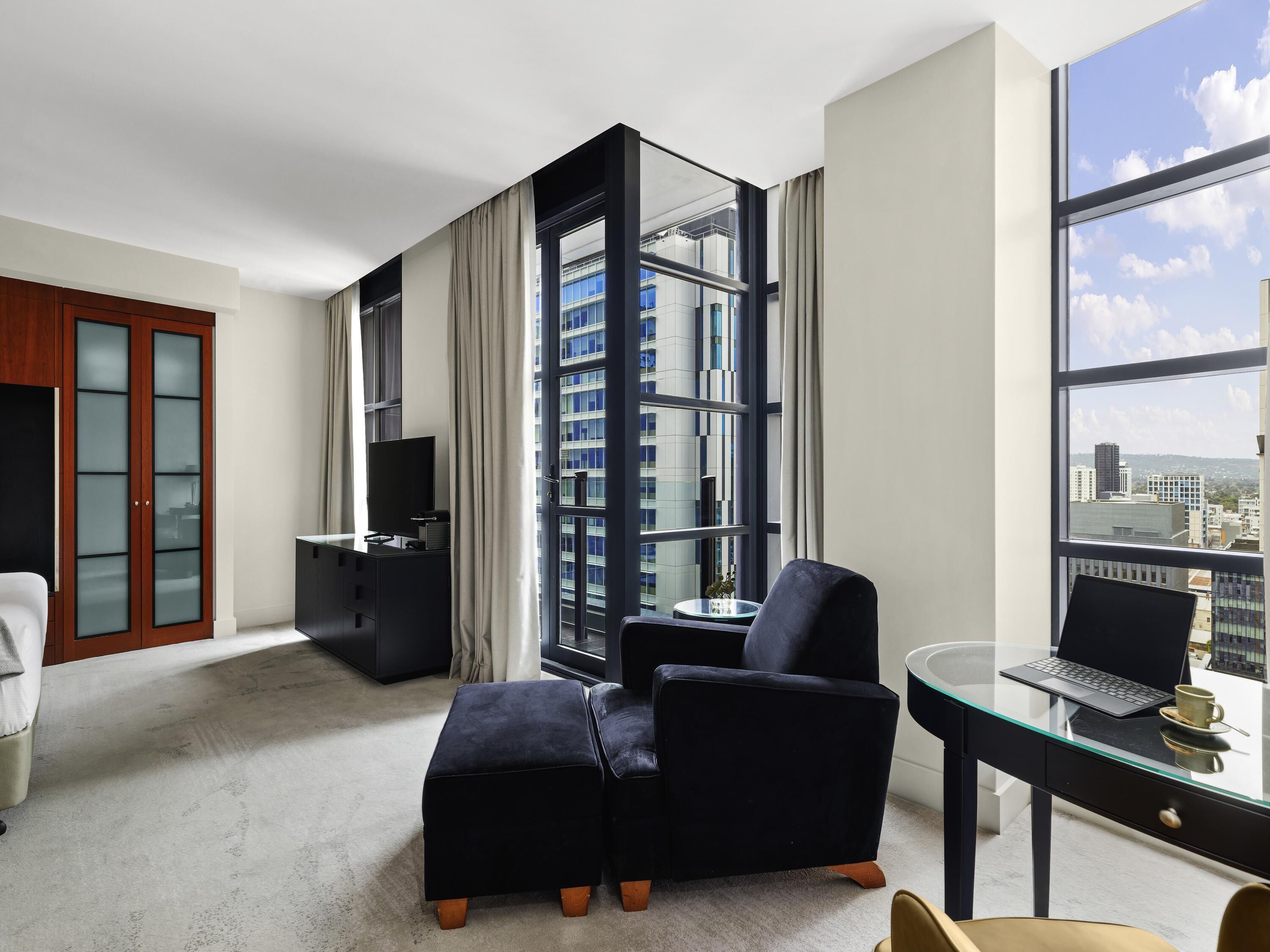 Quarto de hotel moderno com uma janela grande, uma escrivaninha com um laptop, uma poltrona preta e uma televisão.