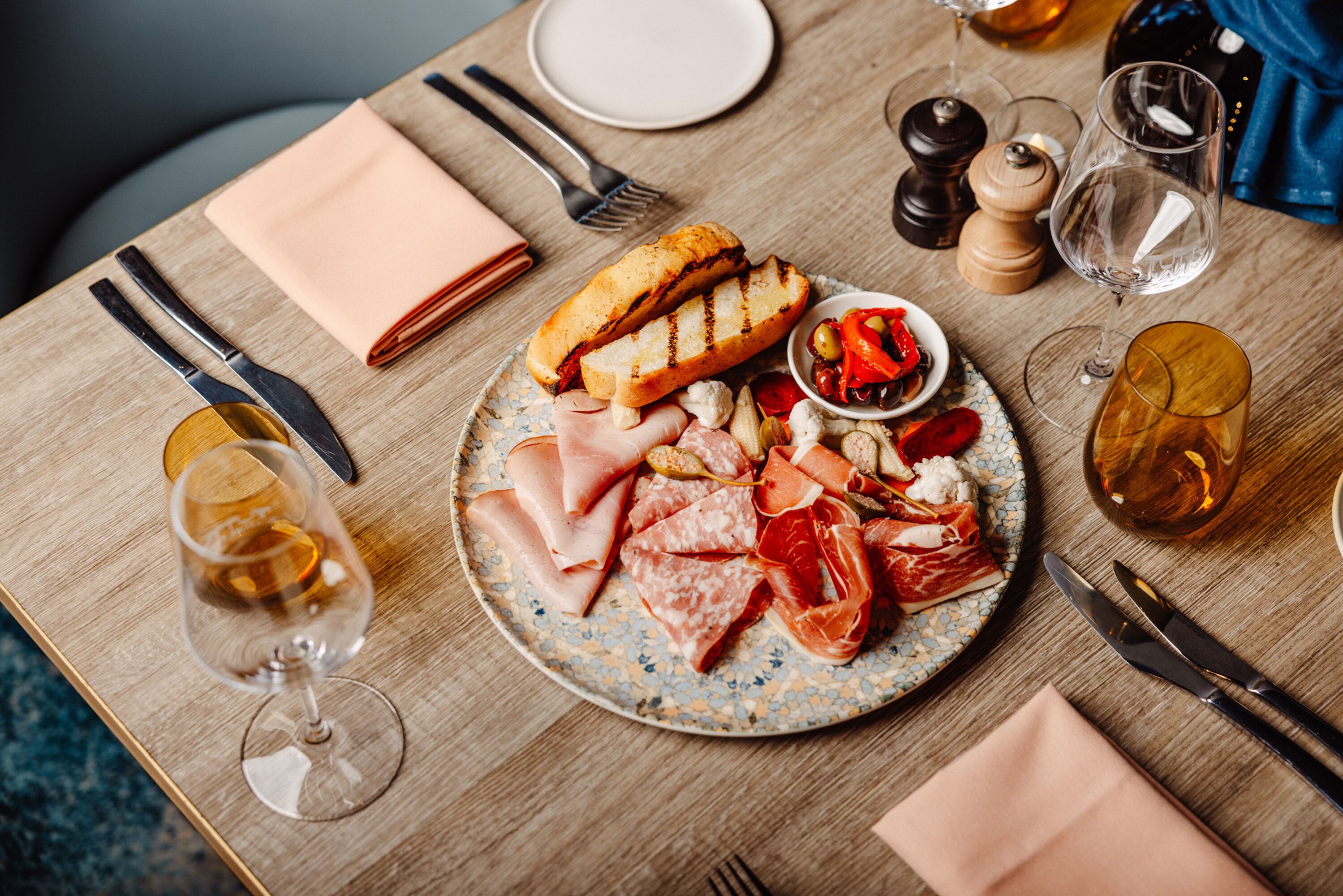 Uma mesa posta com um prato de carnes, pão, azeitonas e uma pequena tigela de pimentões vermelhos, acompanhada de copos de vinho e talheres.