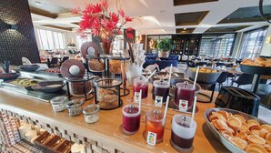 Espaço de buffet com diversas opções de comida e bebidas, uma planta decorativa e uma área de estar ao fundo.