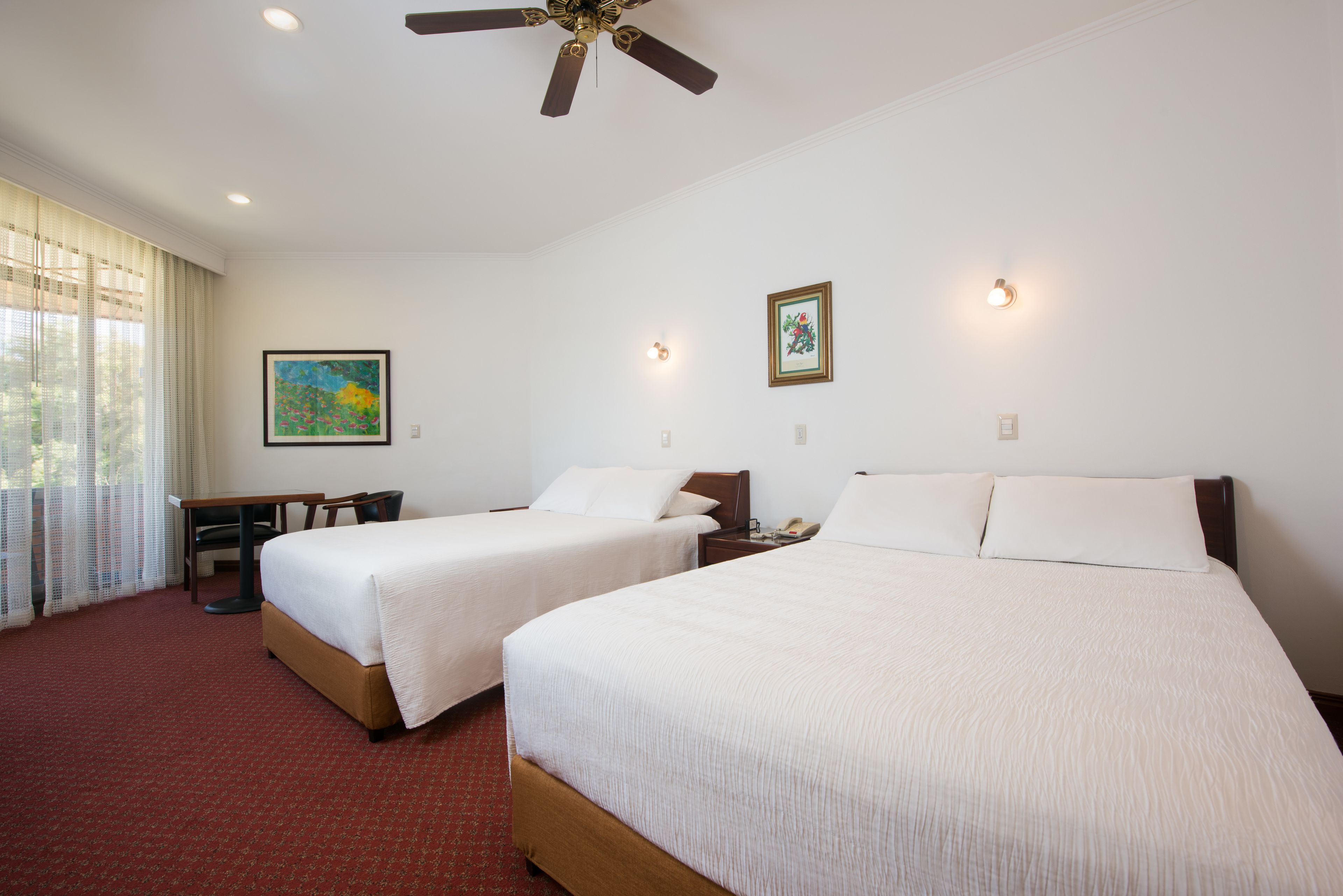 Quarto de hotel com duas camas, uma escrivaninha, uma cadeira, ventoinha no teto e obras de arte nas paredes.