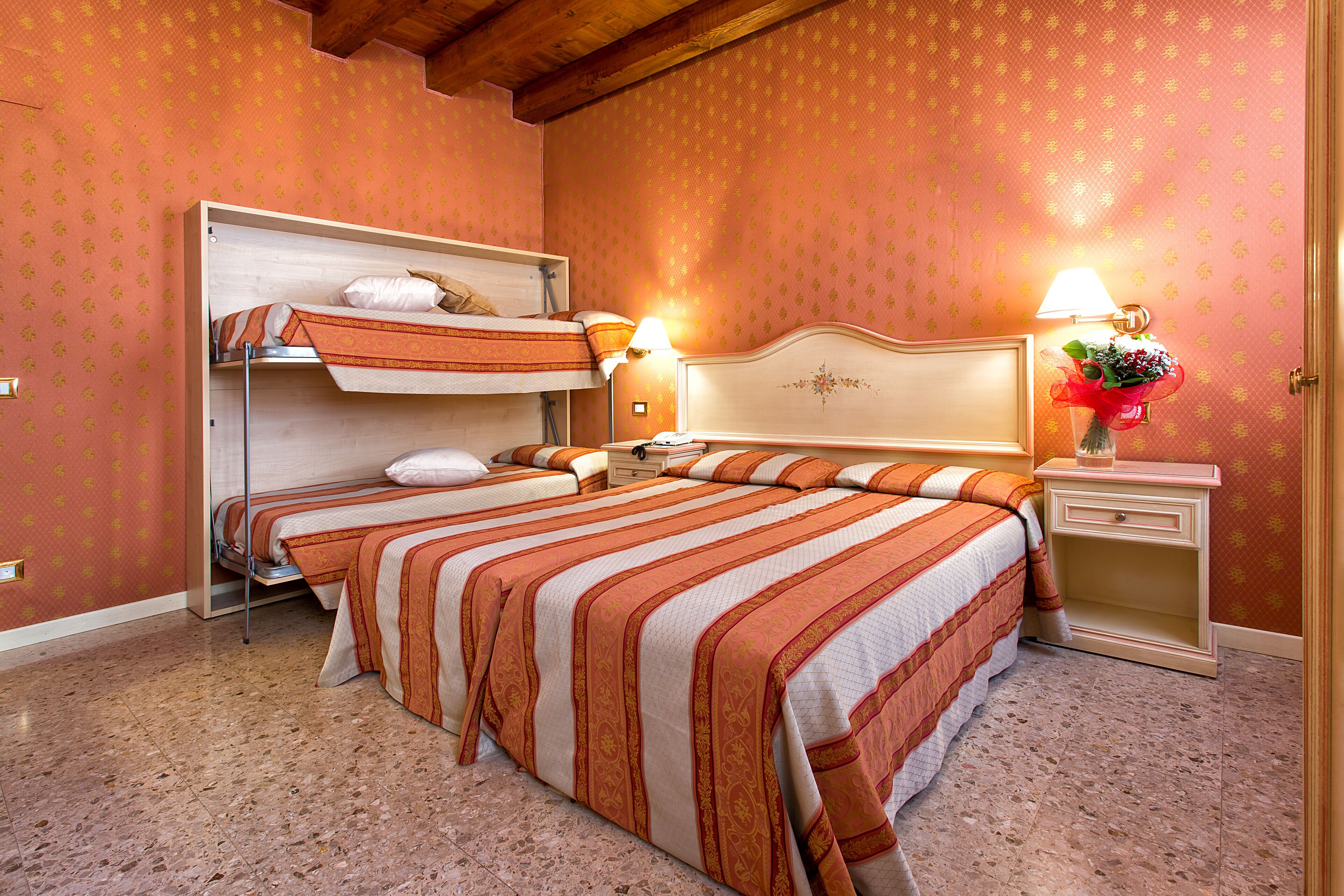 Quarto de hotel com uma cama beliche, duas mesas de cabeceira, um criado-mudo com abajur e um arranjo floral.