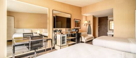 Quarto de hotel com duas camas, uma escrivaninha, um espelho, um pequeno espaço de cozinha e um banheiro.