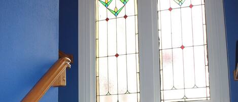 Escadaria com carpete azul e corrimãos de madeira, conduzindo a uma janela com vitrais.