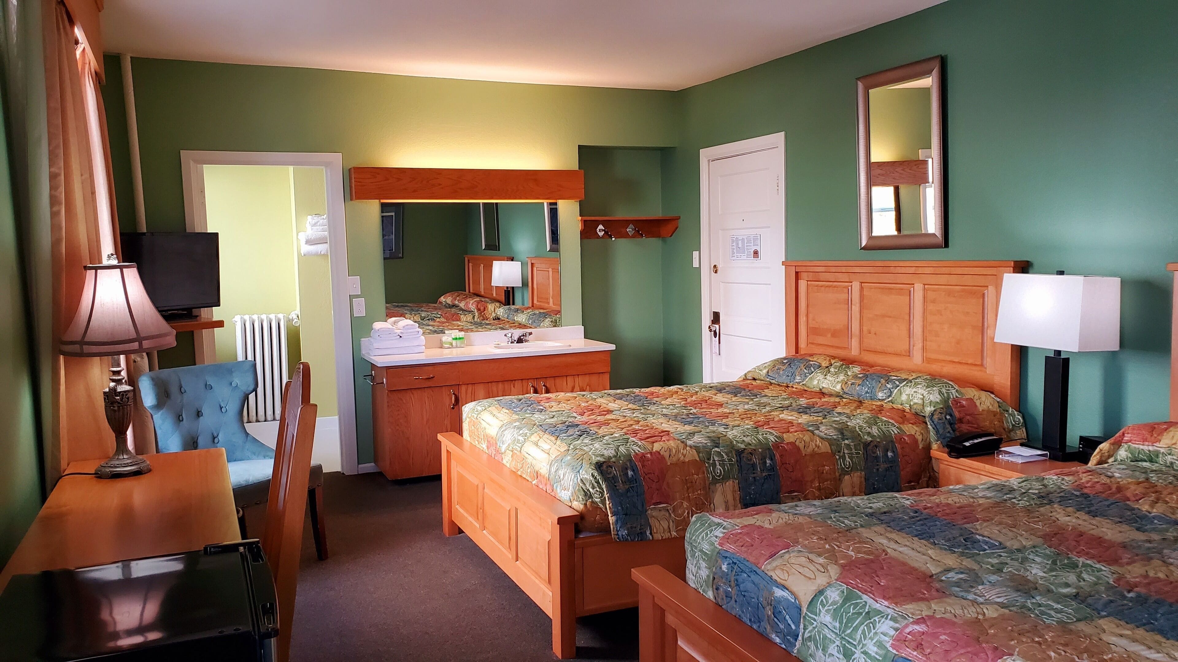 Quarto de hotel com duas camas, cômoda de madeira, uma cadeira, escrivaninha com luminária e televisão.