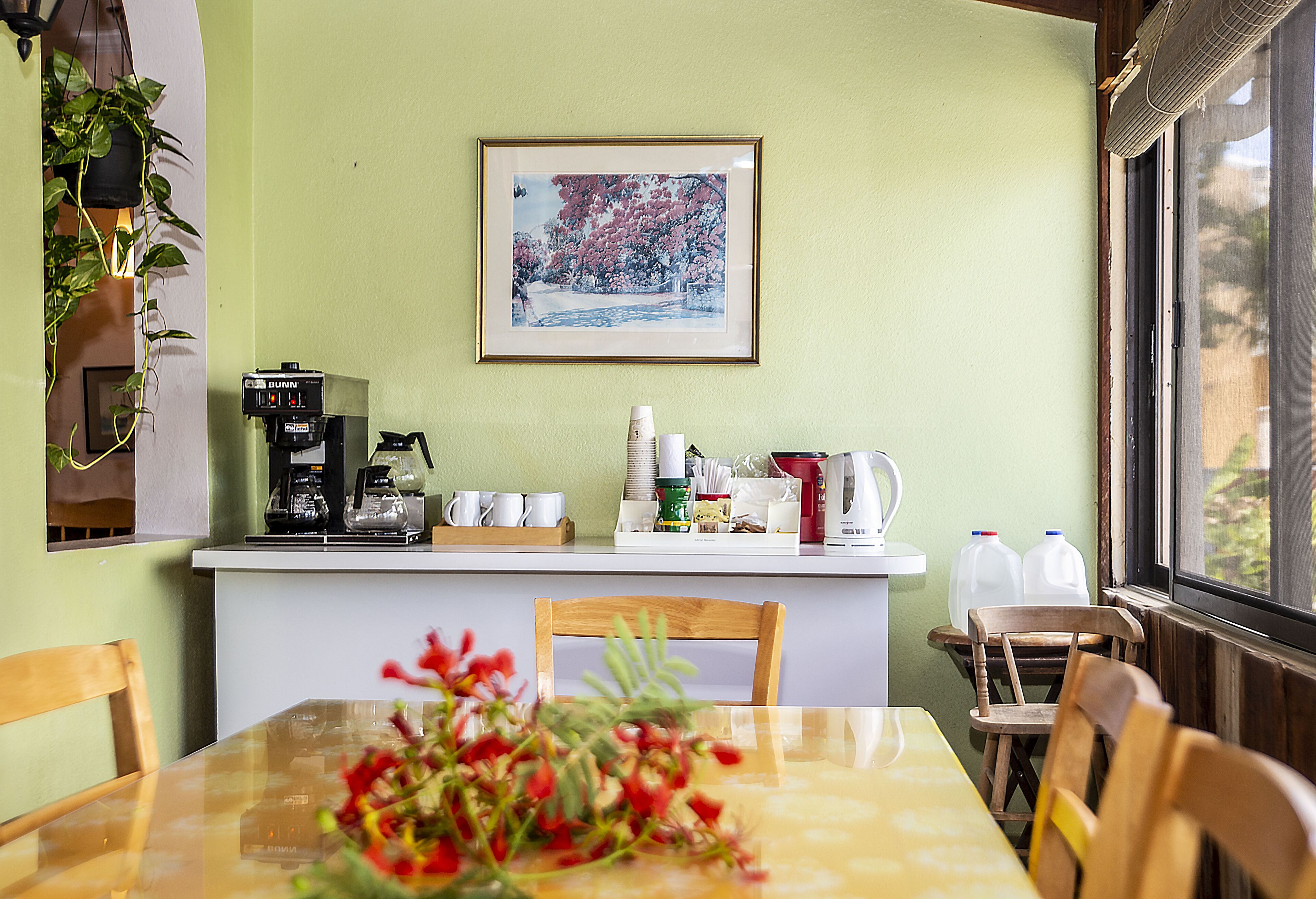 Uma sala de jantar com uma mesa, cadeiras, máquina de café, chaleira e um quadro na parede.