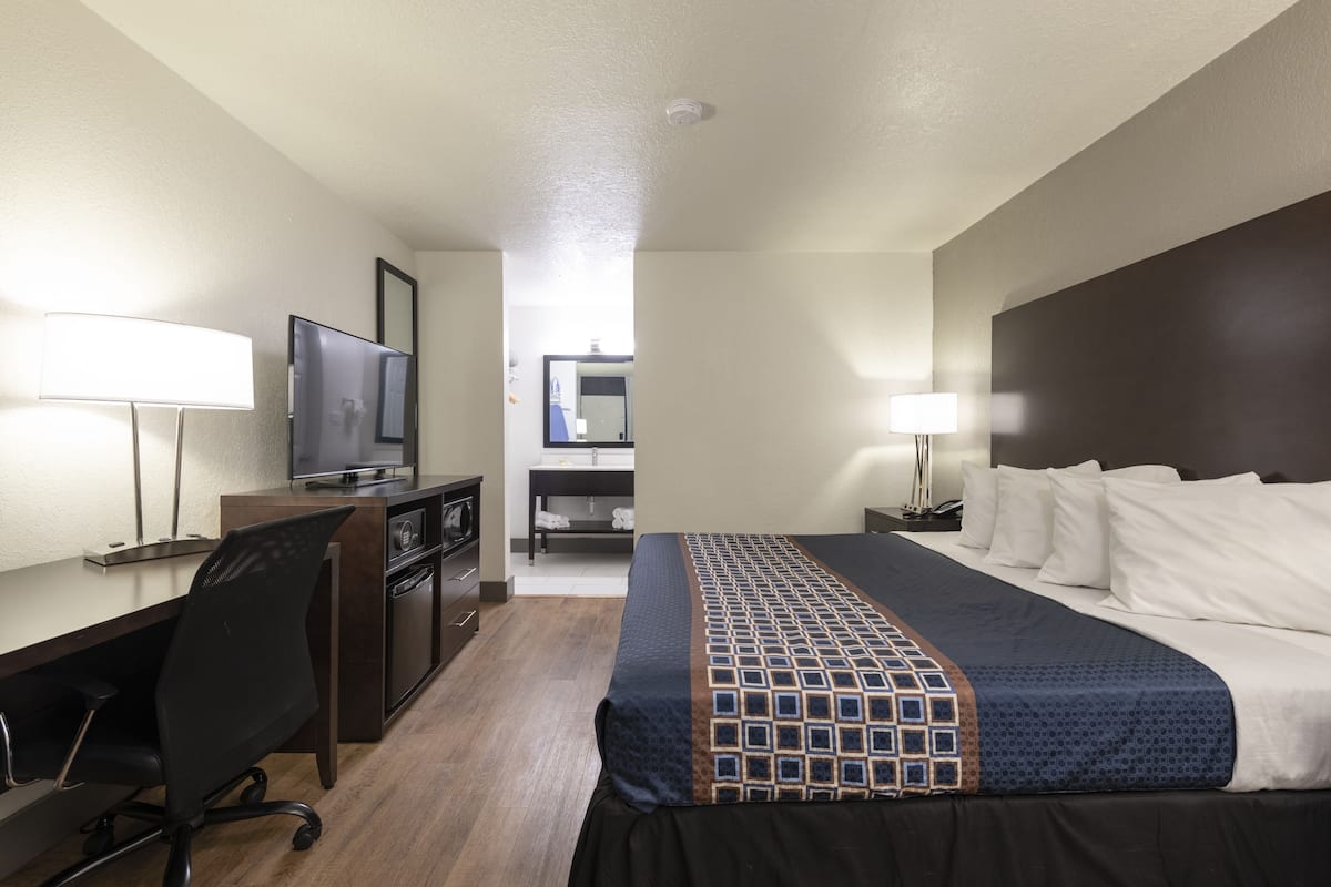 Quarto de hotel com cama, escrivaninha, cadeira, televisão e banheiro ao fundo.