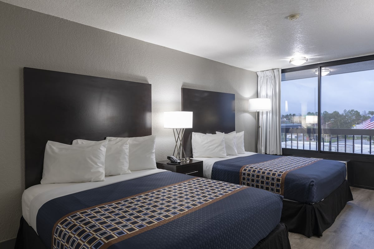 Quarto de hotel com duas camas, uma janela grande com vista e uma mesa de cabeceira com abajur.