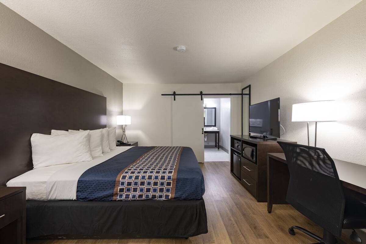 Quarto de hotel com cama, escrivaninha, cadeira, televisão e banheiro visível ao fundo.