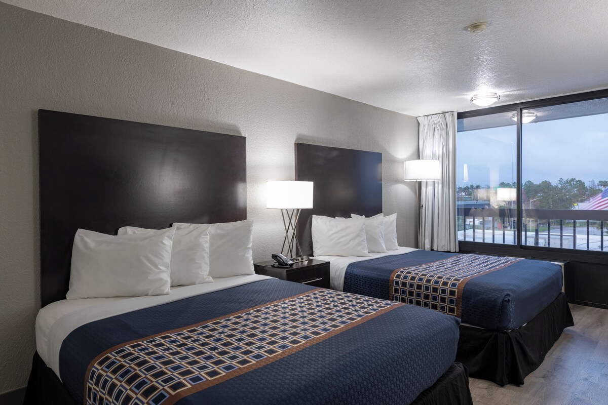 Quarto de hotel com duas camas, uma janela grande com vista e uma mesa de cabeceira com abajur.