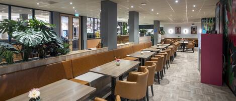 Um restaurante moderno com mesas de madeira compridas e assentos acolchoados, chão em azulejo e amplas janelas que oferecem vistas do exterior.