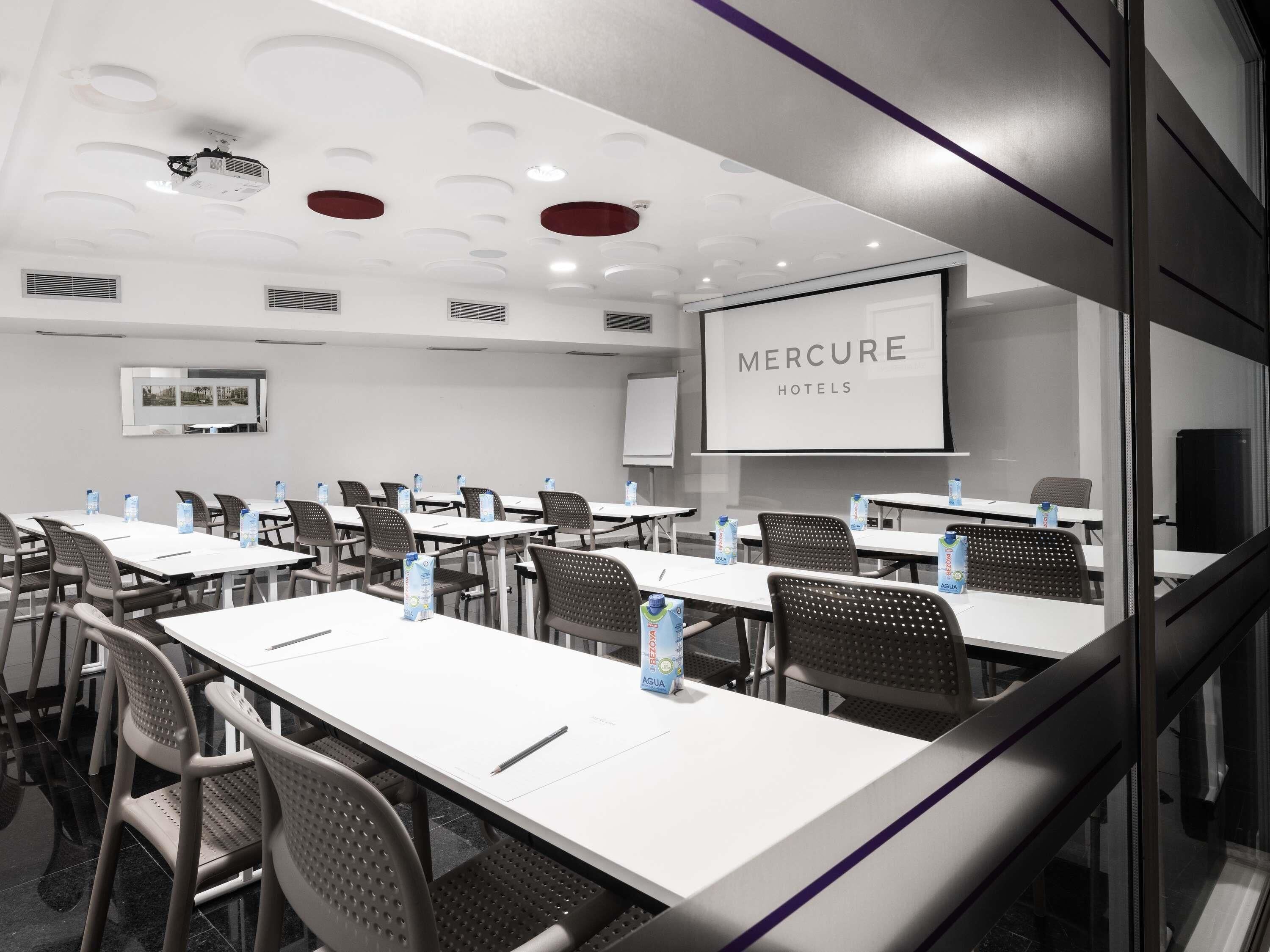 Uma sala de conferências com mesas brancas, cadeiras pretas e uma tela de projeção exibindo 'MERCURE HOTELS'.