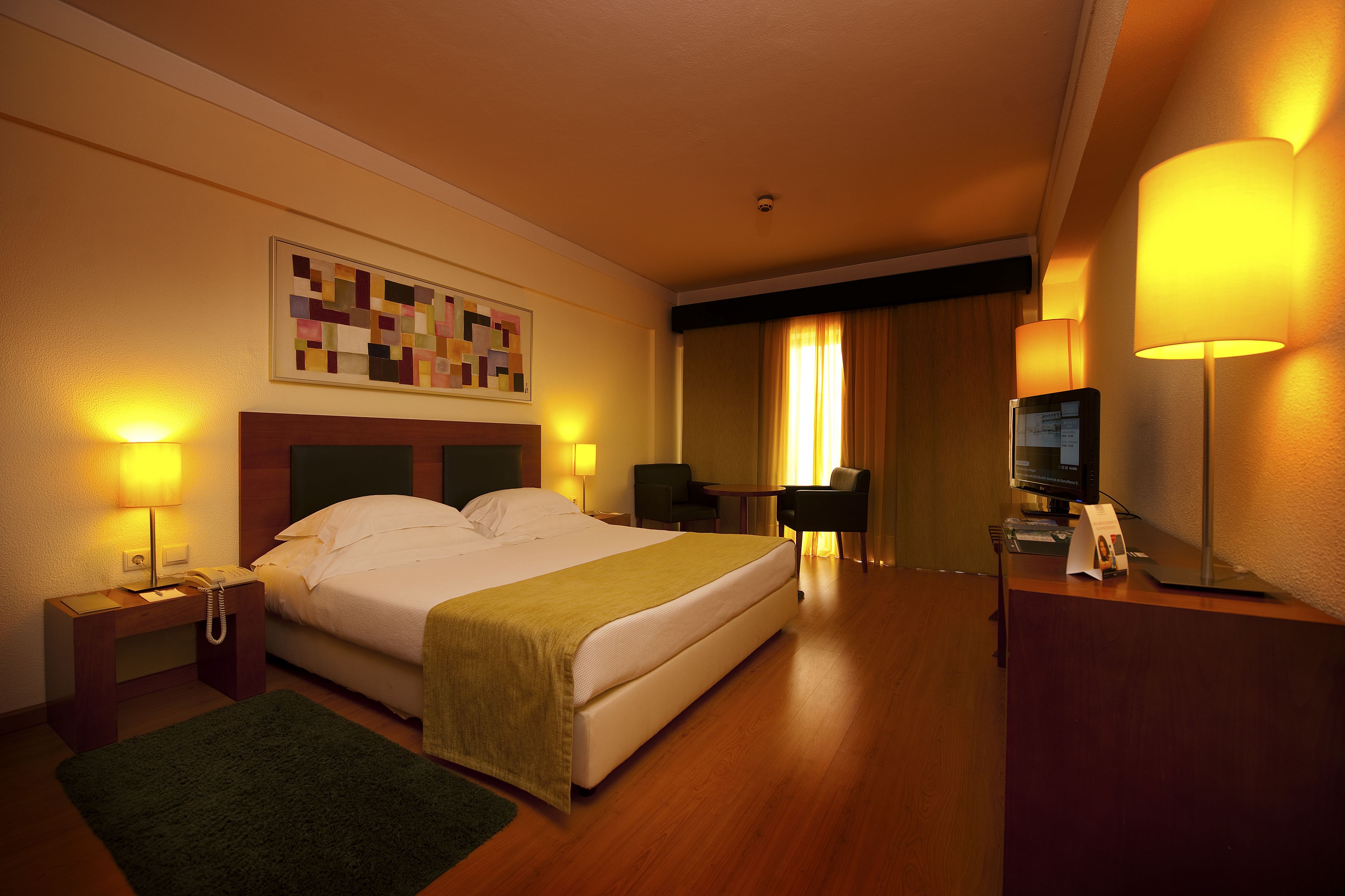 Quarto de hotel com cama, luminárias laterais, secretária com telefone, televisão e um quadro na parede.