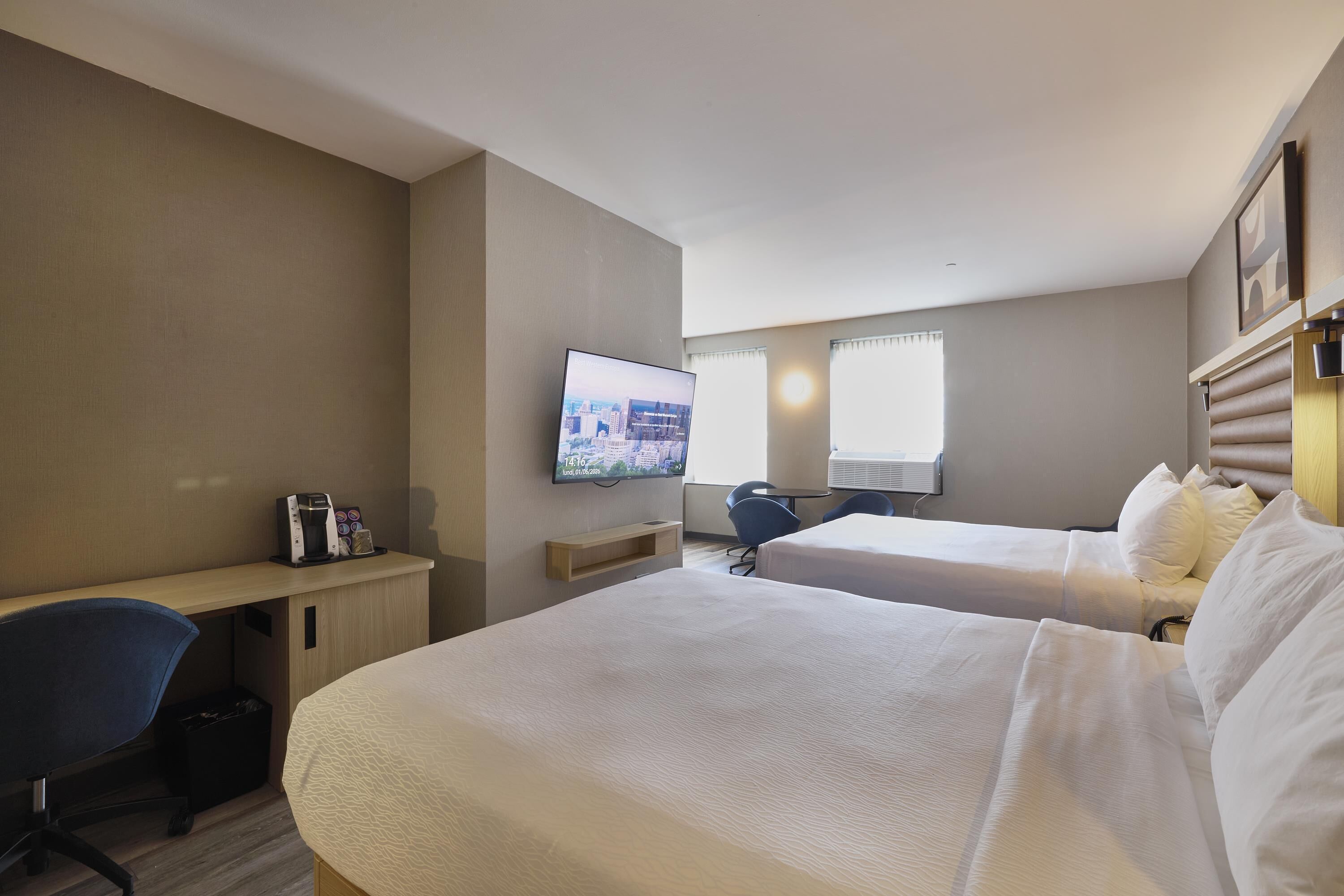 Quarto de hotel com duas camas, uma mesa de trabalho com cafeteira, televisão na parede e uma cadeira.