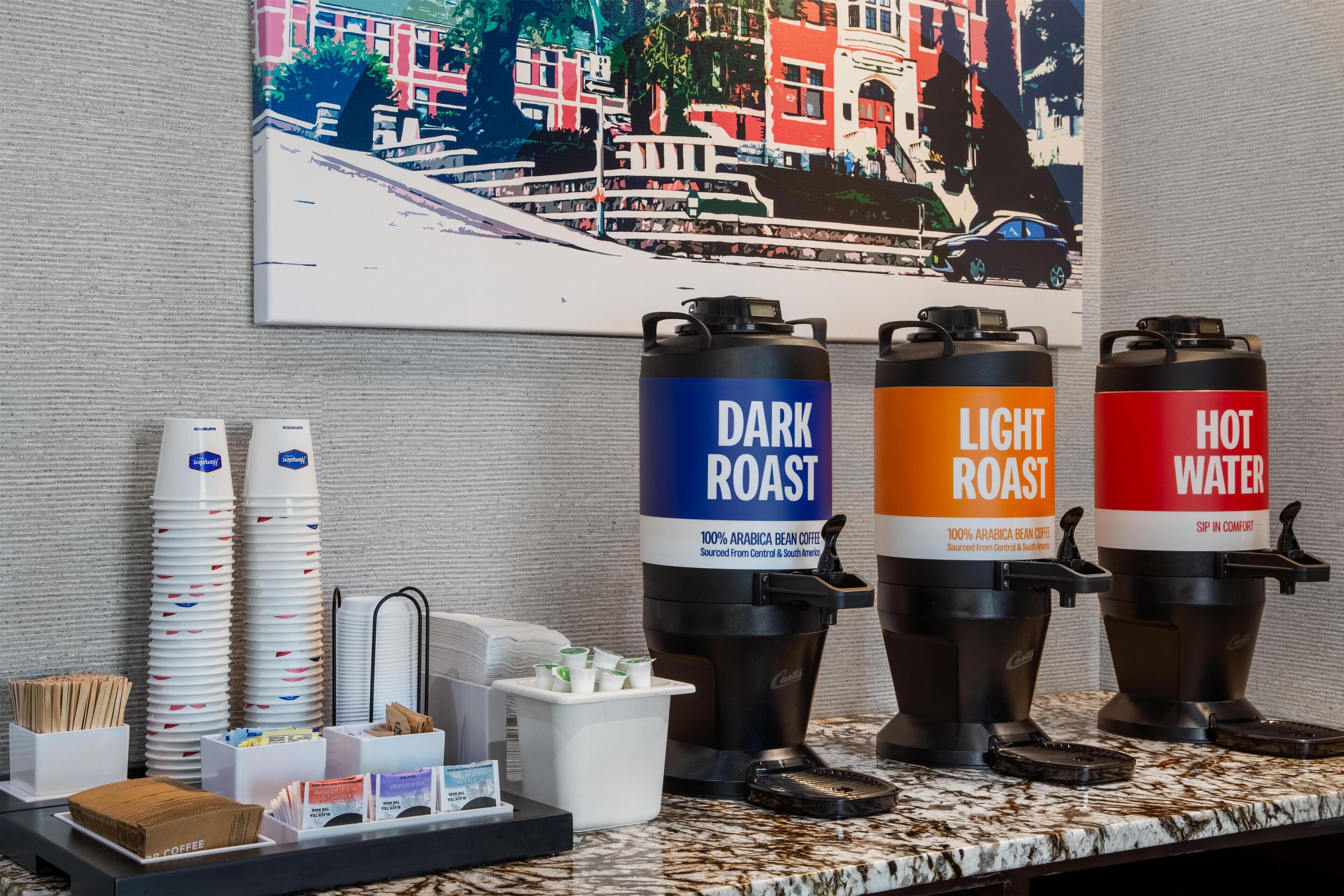 Uma estação de café com três dispensadores de café identificados como 'Dark Roast', 'Light Roast' e 'Água Quente'. Há também pilhas de copos descartáveis e uma bandeja com itens relacionados a café.