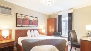 Quarto de hotel com uma cama grande, cabeceira de madeira, duas mesas de cabeceira com luminárias, uma escrivaninha com cadeira, uma janela grande com cortinas e um quadro na parede.