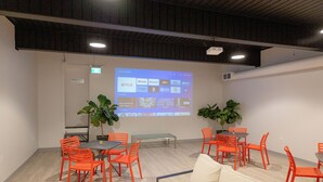 Uma sala de conferência com uma tela grande exibindo a interface de um serviço de streaming, cadeiras laranja, um sofá branco e plantas em vasos.