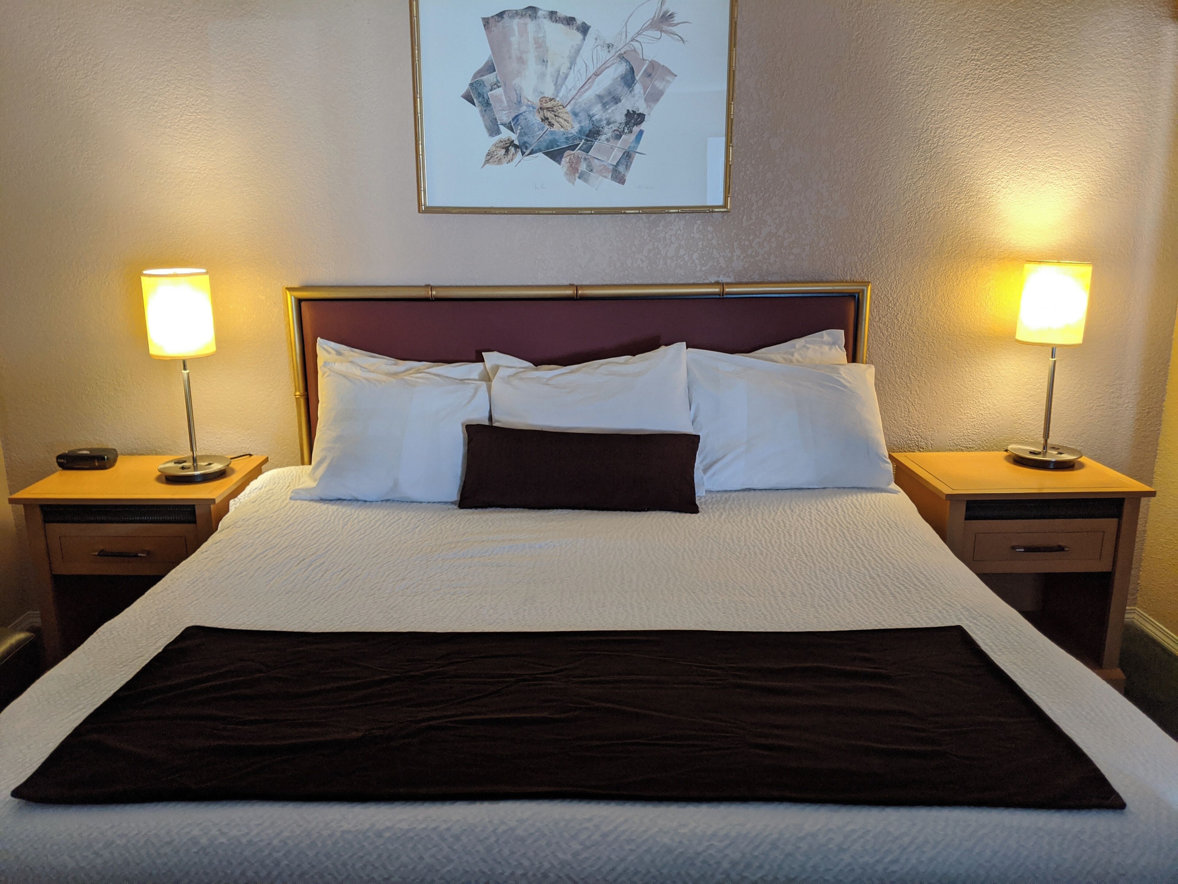 Uma cama bem arrumada com roupa de cama branca e marrom, duas mesas de cabeceira com abajures e uma obra de arte emoldurada na parede.