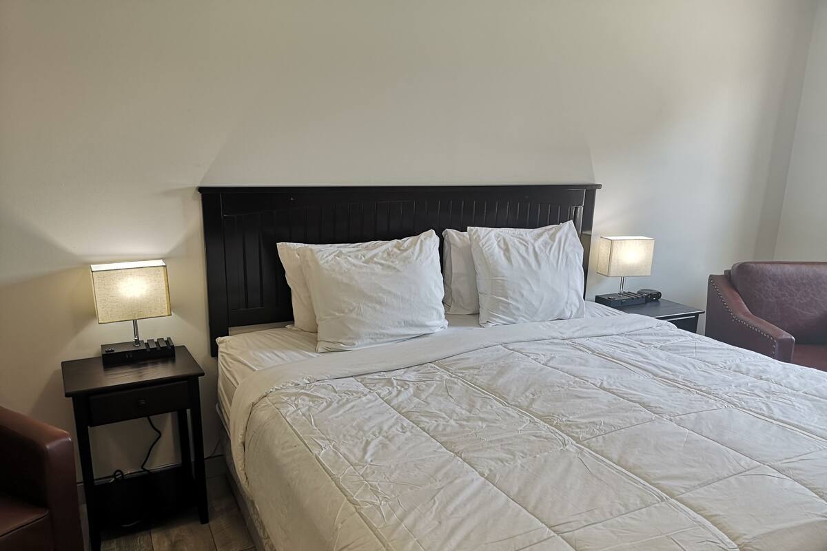 Quarto de hotel com uma cama grande, duas mesas de cabeceira com abajures, uma cadeira e uma cabeceira de madeira escura.