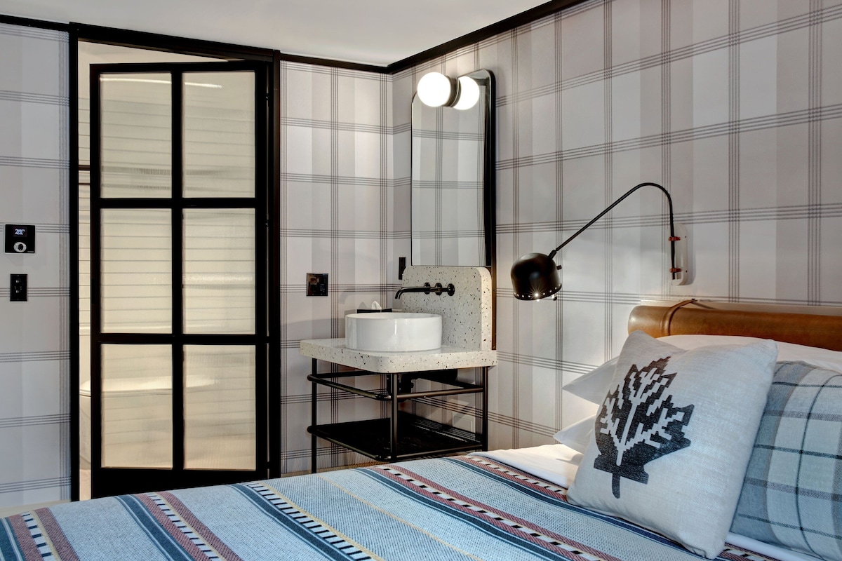 Quarto com cama, mesa de cabeceira com luminária, pia fixada na parede e porta com painéis de vidro jateado.