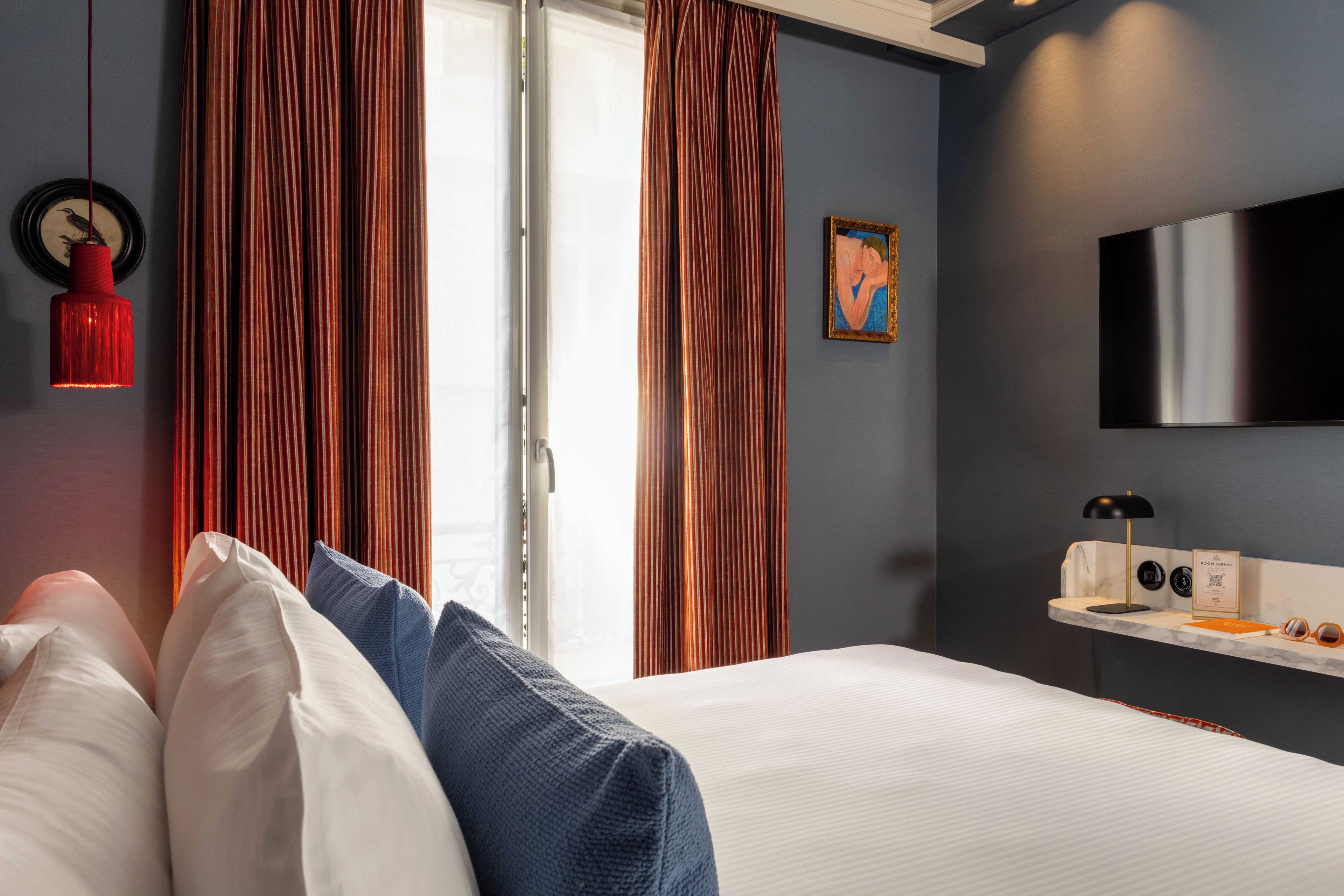 Quarto de hotel com cama, cortinas, relógio, quadro, televisão e escrivaninha com abajur e placa.