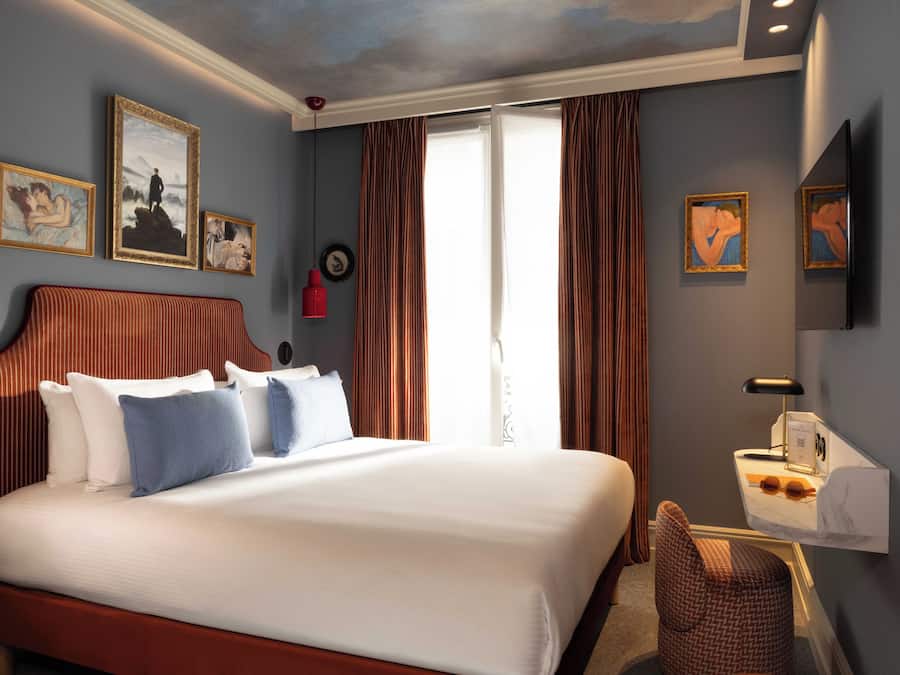 Quarto de hotel com uma cama grande, uma escrivaninha com abajur e obras de arte nas paredes.
