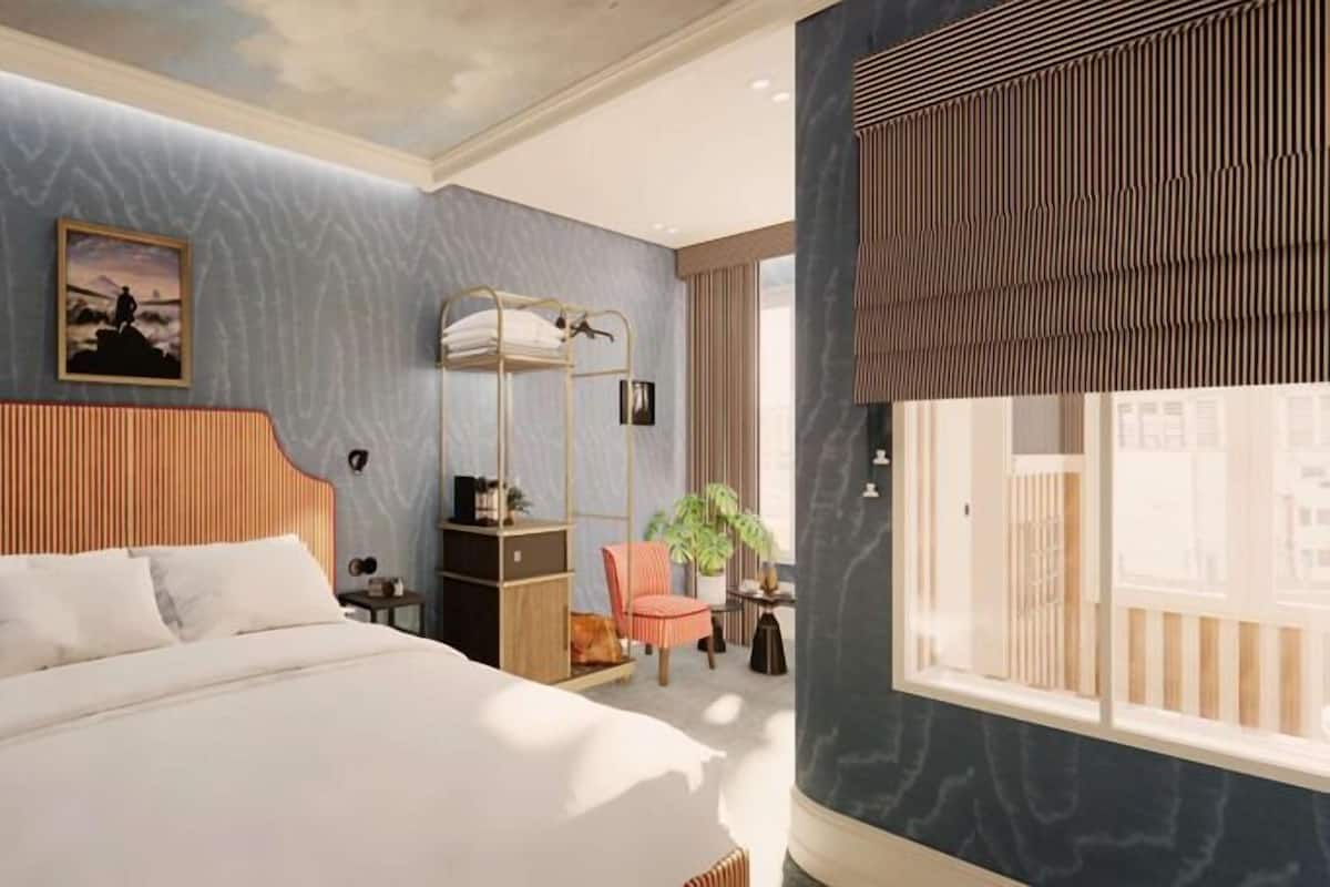 Um quarto com uma cama, mesa de cabeceira, cadeira, janela e uma parede com um quadro.