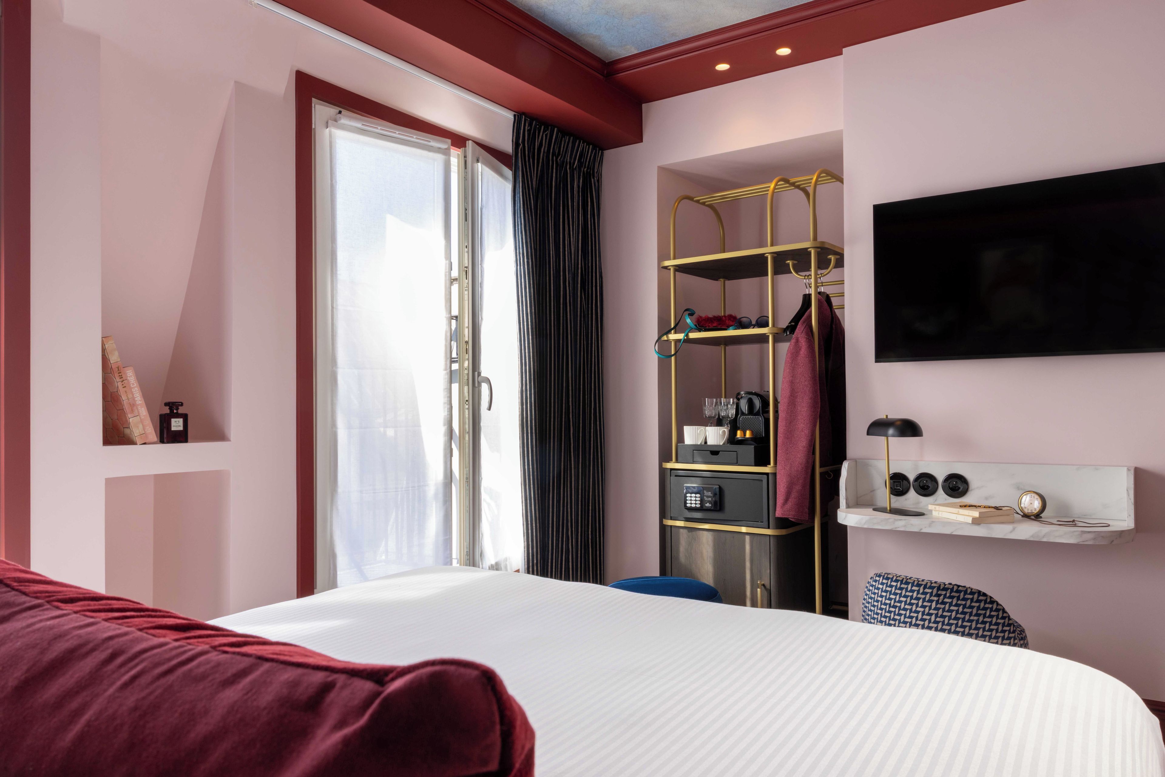 Quarto de hotel com cama, televisão na parede, escrivaninha com computador e janela com cortinas.