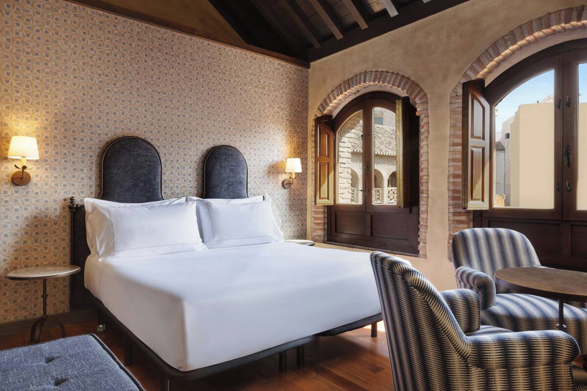 Quarto de hotel bem arrumado, com uma cama grande, duas luminárias fixadas na parede, uma mesa redonda pequena e uma cadeira.