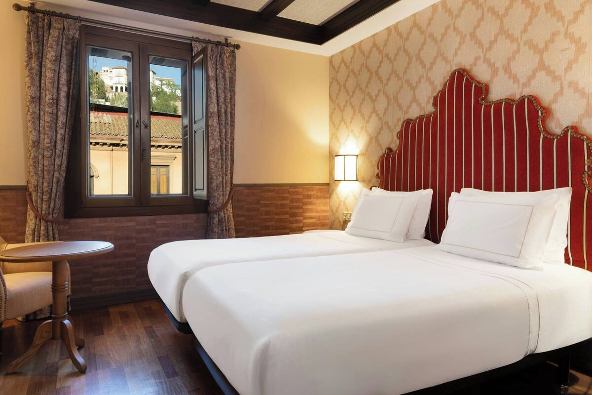 Quarto de hotel com duas camas, uma mesinha redonda, janela com cortinas e parede estampada.