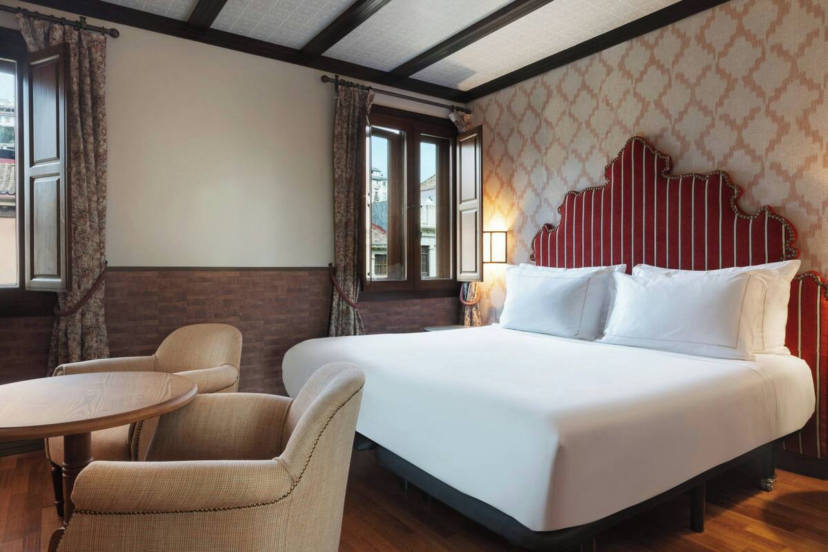 Quarto de hotel com uma cama grande, duas poltronas, uma mesinha e uma janela com cortinas.