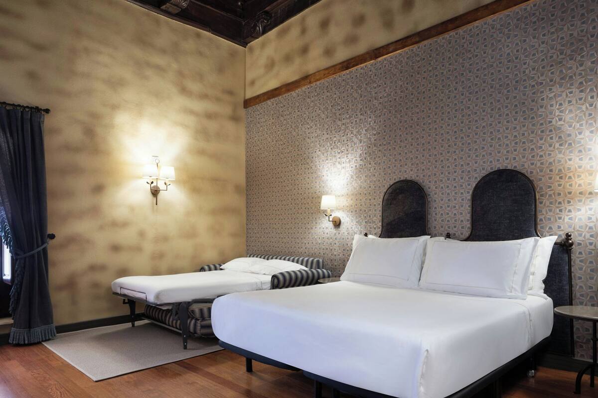 Quarto de hotel com duas camas, piso de madeira, papel de parede estampado e luminárias fixadas na parede.