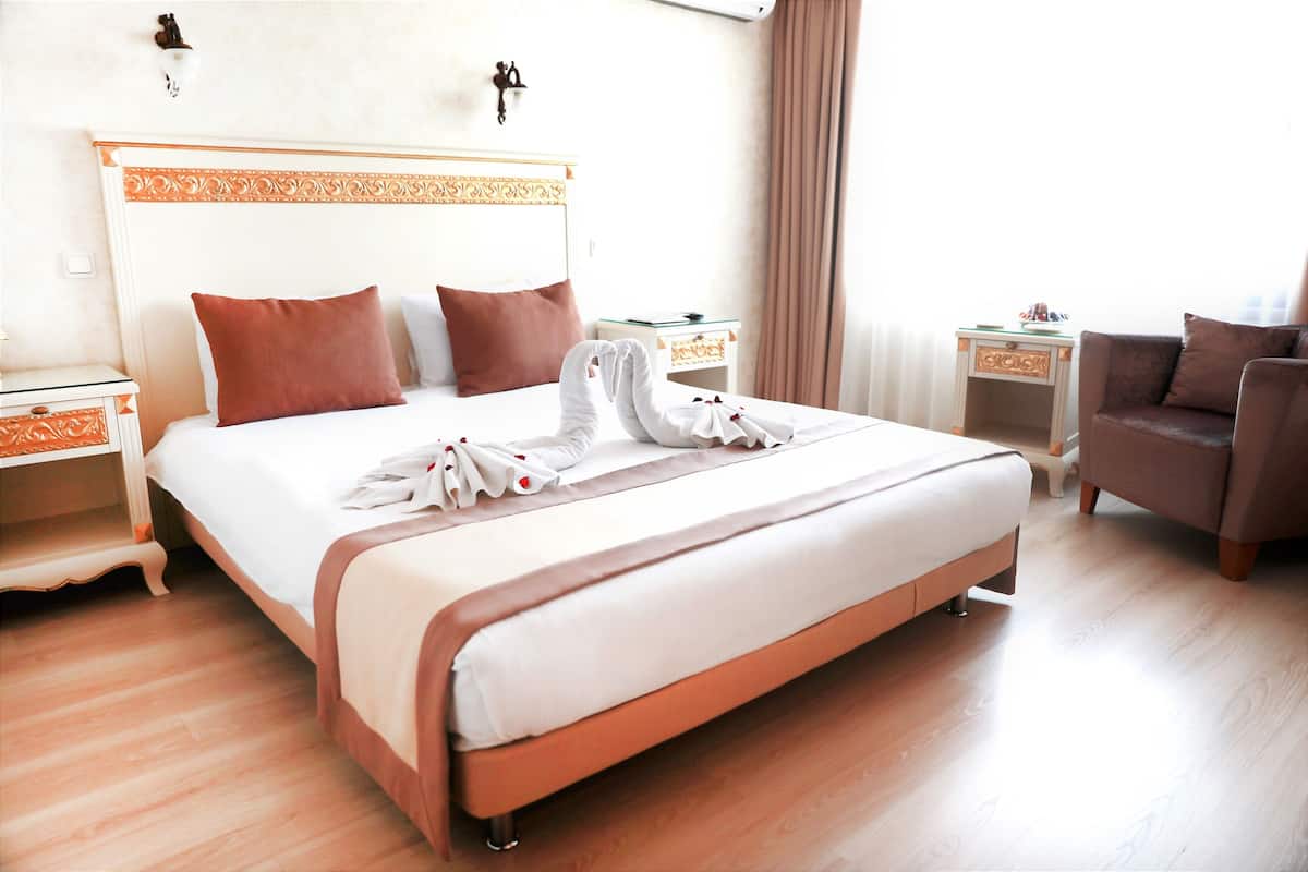 Quarto de hotel com uma cama grande, duas mesas de cabeceira, uma poltrona marrom e uma mesa de vidro.