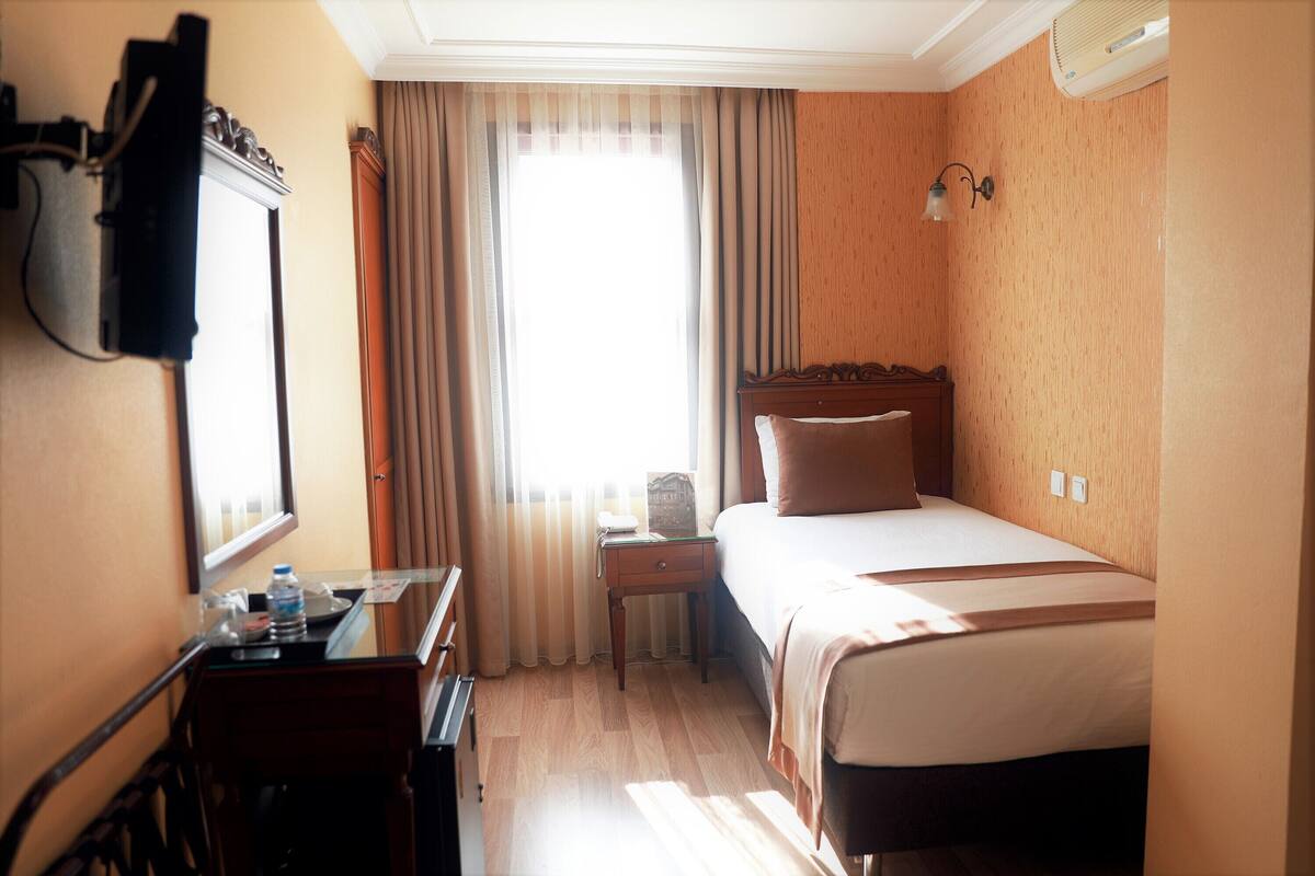 Quarto de hotel com cama, escrivaninha com cadeira, espelho, mesa de cabeceira e janela com cortinas.