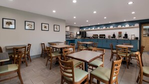 Um restaurante com mesas e cadeiras de madeira, uma área de cozinha e quadros nas paredes.