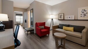 Quarto de hotel com escrivaninha, poltrona vermelha, sofá, luminária de chão e quadros nas paredes.