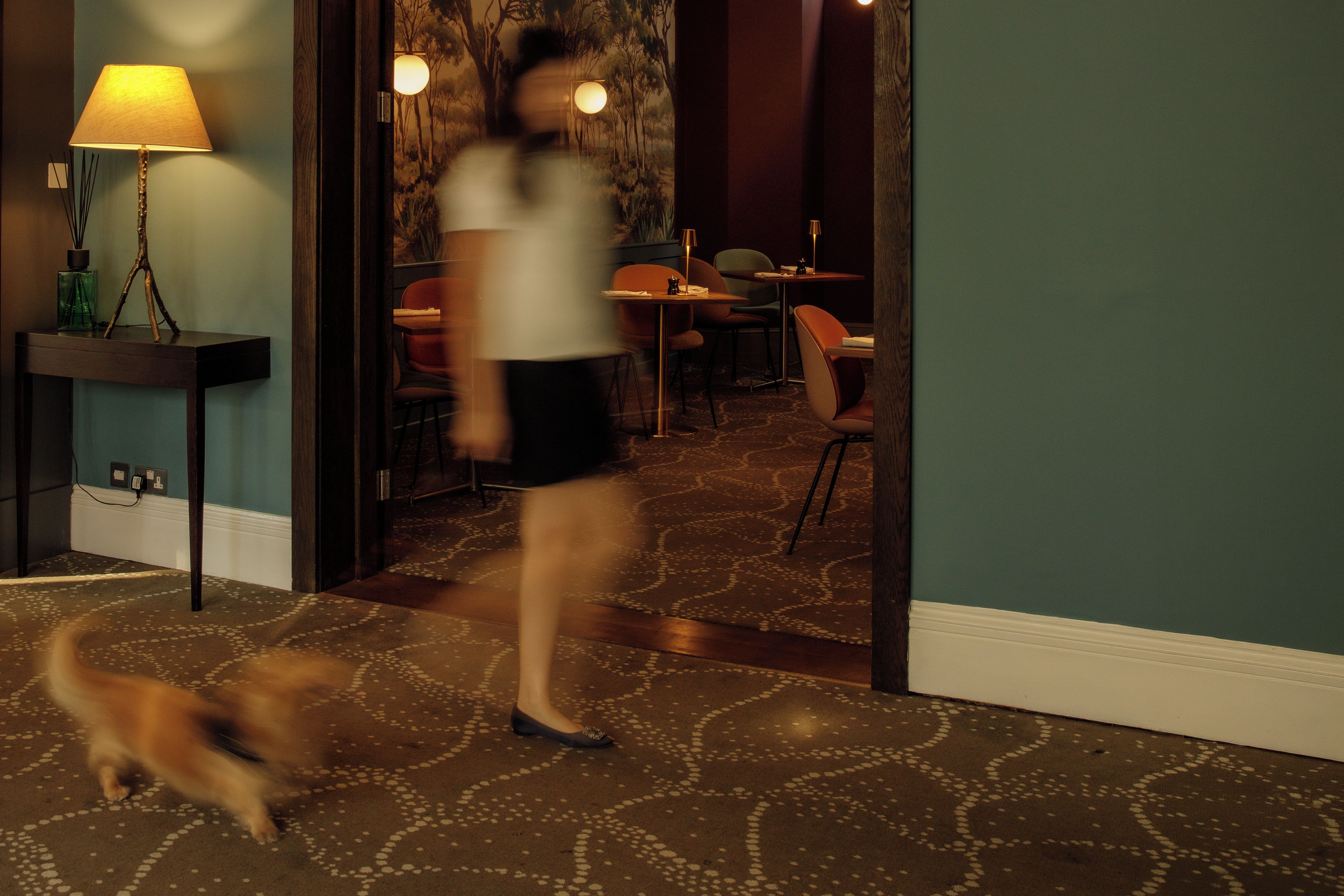 Um hall de hotel com carpete estampado, abajur e área de refeições com cadeiras e mesas.