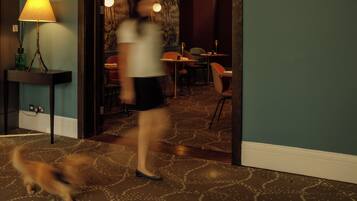 Um hall de hotel com carpete estampado, abajur e área de refeições com cadeiras e mesas.