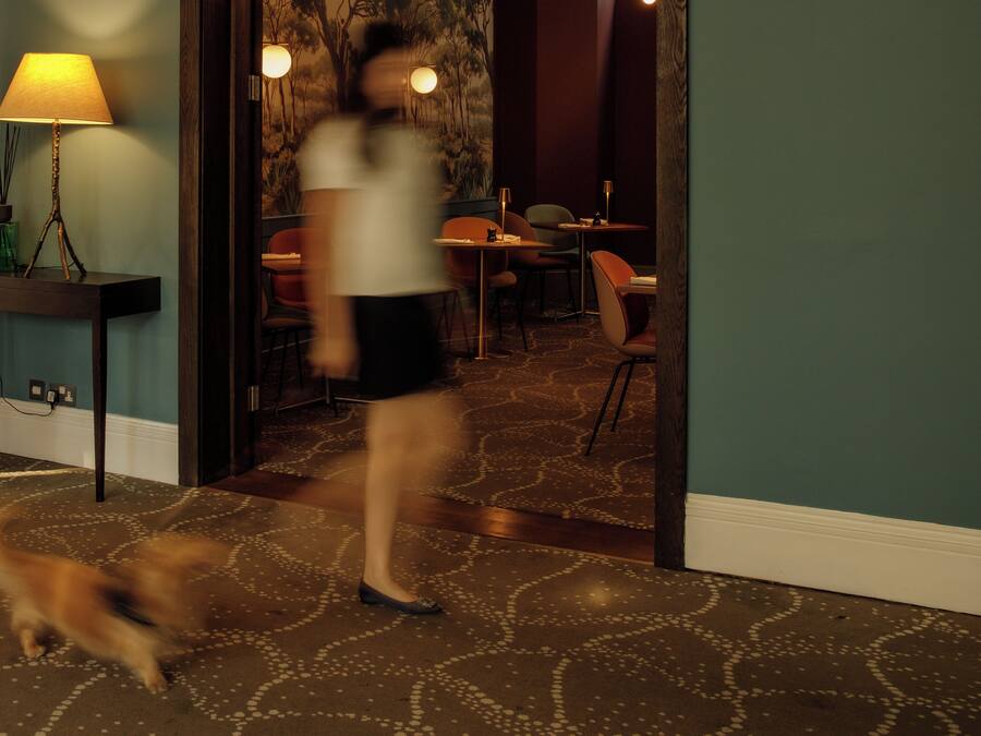 Um hall de hotel com carpete estampado, abajur e área de refeições com cadeiras e mesas.