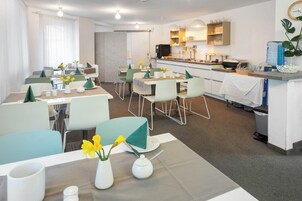 Espaço de refeições moderno em hotel, com mesas preparadas para o pequeno-almoço, zona de cozinha e dispensador de água.