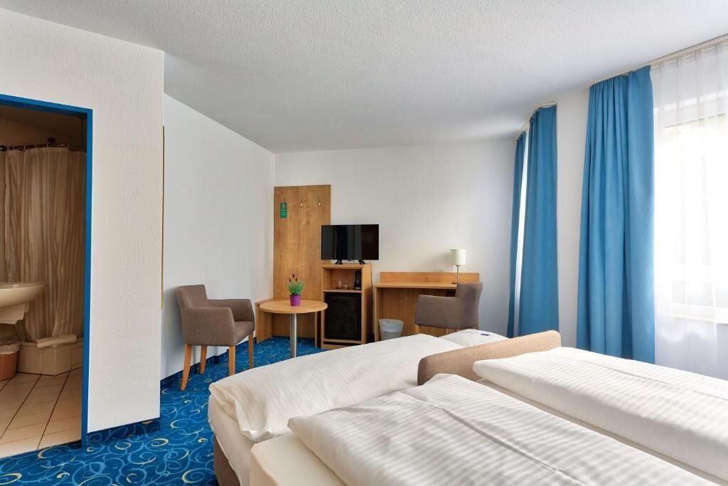 Quarto de hotel com duas camas, uma escrivaninha, uma cadeira, televisão e banheiro com chuveiro.