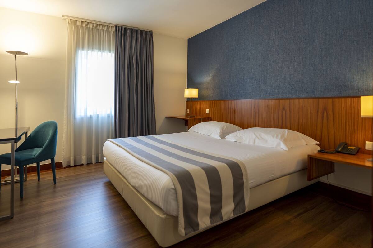 Quarto de hotel com cama, escrivaninha, cadeira, luminária e telefone.