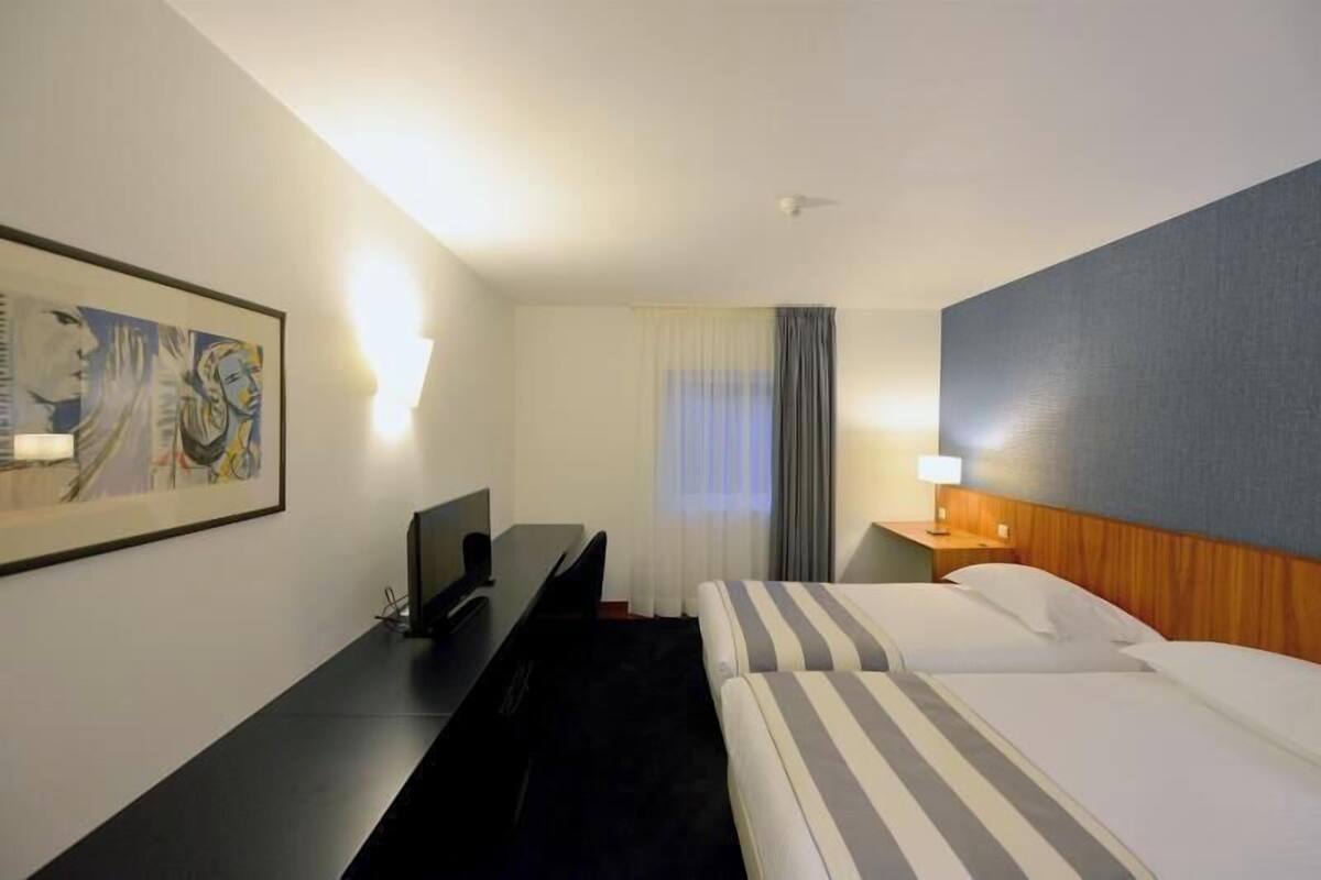 Quarto de hotel com cama, escrivaninha, cadeira, televisão, quadro na parede e janela com cortinas.
