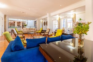 Uma sala ampla com um sofá azul grande, poltronas amarelas e verdes, uma mesa de centro de vidro e um vaso com flores.
