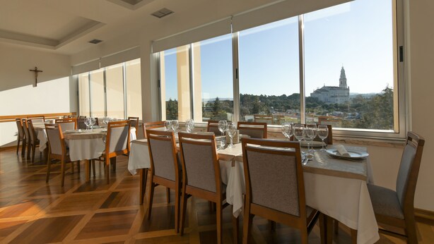 Uma sala de jantar com mesas postas para uma refeição, grandes janelas com vista para um edifício distante e uma cruz na parede.